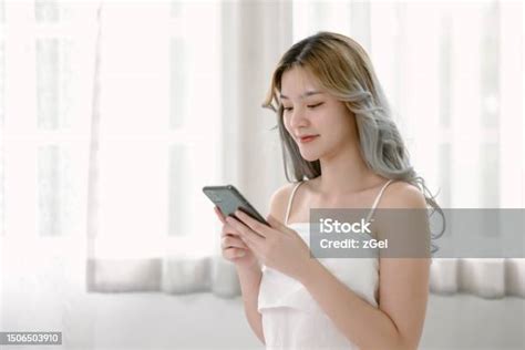 아름 다운 사랑스러운 젊은 아시아 여자는 서서 문자 메시지를 입력하고 휴대 전화로 소셜 미디어를 채팅하고 집 거실에서 온라인 커뮤니케이션을 즐긴다 기술 커뮤니케이션 라이프