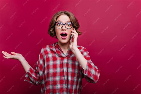 Удивленная молодая женщина с большими темными глазами разговаривает по телефону на бордовой