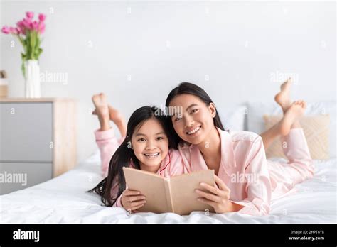 Cheerful Japanese Teenage Girl In Pajama And Young Female Lie On Bed And Read Book In Bedroom