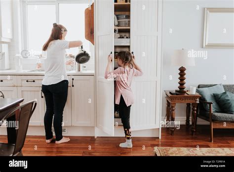 Mother Pouring Hot Water Girl Looking Into Cabinet In Kitchen Stock