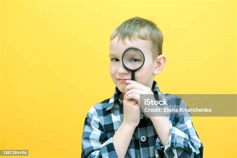 캐주얼 한 옷을 입은 긍정적 인 호기심 많은 여학생이 노란색 배경에 돋보기를 통해 카메라를 봅니다 과학에 대한 스톡 사진 및