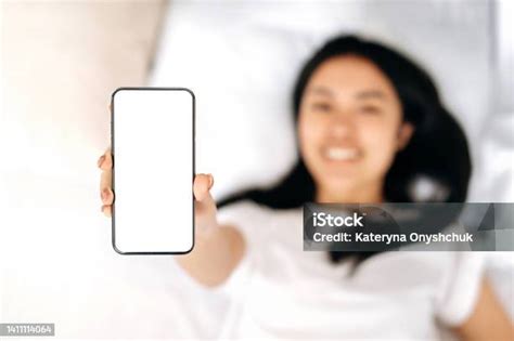 광고 또는 프리젠 테이션을위한 모형 개념 집중이 잘되지 않은 아시아 소녀는 침대에 누워 웃고 빈 흰색 화면이있는 스마트 폰을 보여 주며 공간을 복사합니다 무선 기술 20