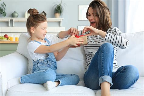 어린 소녀는 엄마와 함께 소파에 앉아있는 동안 흥분 된 부모에게 생일 선물을 만들고 미소 짓는 어린 아이는 행복한 어머니에게 선물 상자를 제공합니다 2명에 대한 스톡 사진