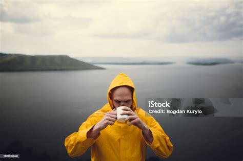 금속 머그잔을 들고 호수를 볼 수있는 비오는 날 절벽에 서있는 노란색 비옷을 입은 힙스터 여행자 방랑자와 여행 잔인한 수염 남자 하이킹 대기 모멘트 감정에 대한 스톡 사진