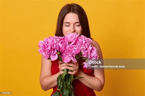 노란 스튜디오 벽에 분홍색 모란의 꽃다발을 들고 드레스를 입고 꽃 냄새를 맡고 눈을 감고 있는 매력적인 소녀의 초상화를 닫습니다