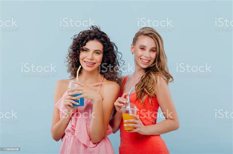 곱슬 머리와 유행 두 쾌활한 여자 주스와 플라스틱 컵을 잡고 파란색 배경에 고립 된 카메라 긍정적 인 얼굴 감정을보고 복사 공간 여자만에 대한 스톡 사진 및 기타 이미지
