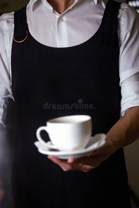 Barista In A White Shirt And Black Apron Holds A Cup Of Coffee Stock