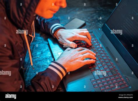 A Hooded Hacker Is Typing On A Laptop Keyboard In A Dark Room Under A