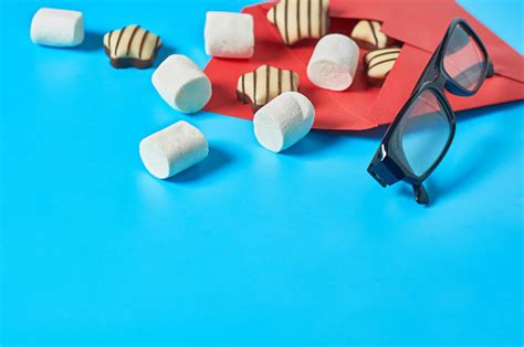 흩어져있는 과자 줄무늬 사탕 검은 색 플라스틱 프레임의 광학 안경 근처의 원통형 마시멜로가있는 빨간 종이 봉투가 파란색 책상