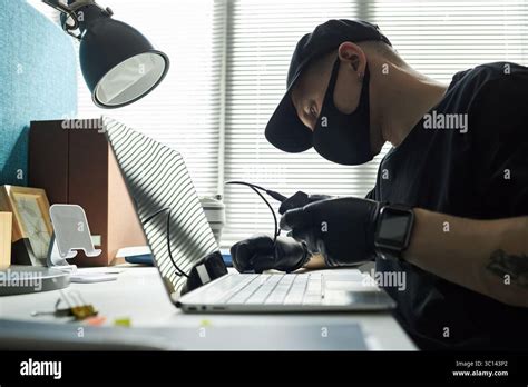 Male Hacker Wearing Black Cap And Face Mask Sitting At Desk In Office Breaking Into Corporate