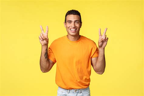 오렌지 색 티셔츠에 잘 생긴 즐거운 젊은 남성 미소 낙관적 평화 제스처 또는 따옴표 기호를 노란색 배경을 서 긍정적 인 표현과 낙관 노란색 배경을 서 건강한 생활방식에 대한