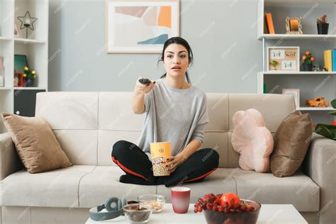 Tv 리모컨을 들고 팝콘 양동이를 들고 거실에 있는 커피 테이블 뒤에 소파에 앉아 있는 혼란스러운 어린 소녀 무료 사진