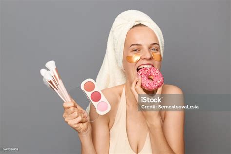 회색 배경 위에 격리된 머리에 상의와 수건을 쓰고 브러시와 아이섀도를 들고 맛있는 도넛을 물고 있는 흥분된 배고픈 웃는 여자의
