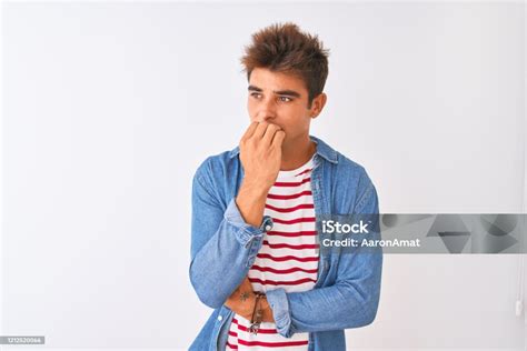 줄무늬 티셔츠와 데님 셔츠를 입은 젊은 잘생긴 남자는 고립 된 흰색 배경 위에 스트레스와 긴장한 모습으로 입에 물린 손톱을 물고 있습니다 불안 문제 T 셔츠에 대한 스톡
