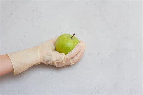고무 장갑을 낀 손에는 녹색 사과 열매가 있습니다 녹색 사과는 밝은 회색과 흰색 현대 콘크리트 배경에 흰색 일회용 고무 라텍스