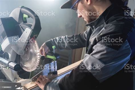 젊은 갈색 머리 남자가 직업에 의해 회색 재킷에 모자를 입고 목수는 워크샵의 워크 벤치 테이블에 원형 톱으로 나무 보드를 잘라