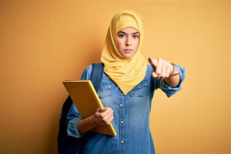 무슬림 히잡과 배낭을 입고 젊은 학생 소녀는 카메라와 당신에 손가락으로 가리키는 노란색 배경을 통해 책을 들고 손 기호 앞에서 긍정적이고 자신감 제스처 가리키기에 대한 스톡