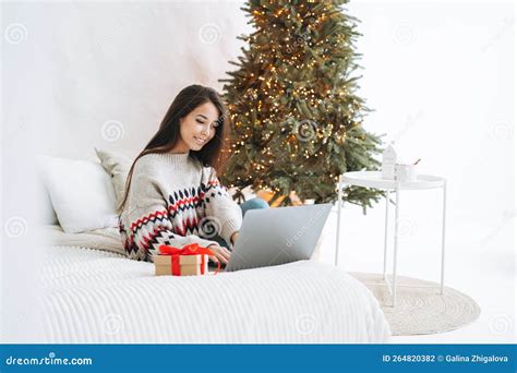 Young Asian Woman In Cozy Sweater Using Laptop On Bed In Room With
