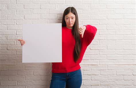 성 난 얼굴 아래로 엄지 싫어 보여주는 음수 부호 거부 개념 배너를 들고 벽돌 벽을 통해 젊은 중국 여자 개념에 대한 스톡 사진