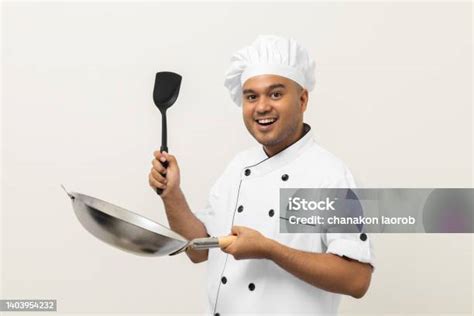 유니폼을 입은 젊은 잘 생긴 아시아 남자 요리사 터너와 철제 프라이팬 기구 부엌에서 요리하는 것은 고립 된 배경에서 다양한 제스처를 취합니다 인도 남자 직업 요리사