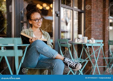 Hispanic Woman In Eyeglasses Smiling And Reading Book At Cafe Stock