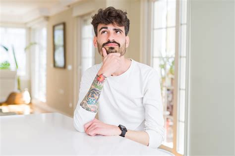 젊은 남자가 턱에 손으로 흰색 테이블에 앉아 캐주얼 셔츠를 입고 질문 잠겨있는 표현에 대해 생각 합니다 사려 깊은 얼굴로 웃 고 있습니다 의심의 개념입니다 남자에 대한 스톡