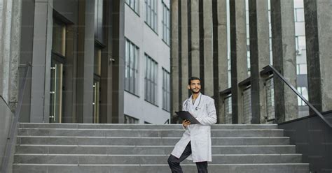 젊은 잘 생긴 숙련 된 남성 아랍 건강 관리 노동자의 전체 길이 폴더 클립 보드에 메모를 작성 현대 건물의 계단에 서 야외에서 의료 기록을 작성하는 전문 인도 의사