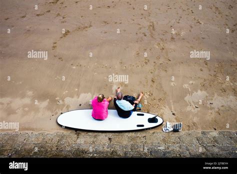 Looking Down On An Older Couple Sitting On A Surfboard On A Sandy Beach