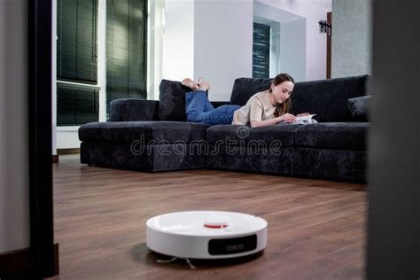 Woman Lying On Sofa And Reading A Book While Robot Vacuum Cleaner Doing Housework Stock Image