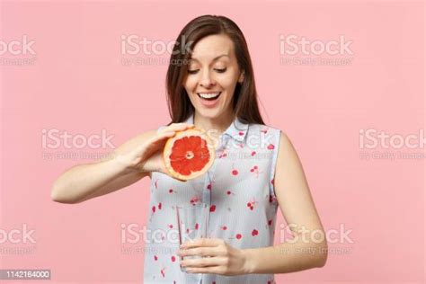 분홍색 파스텔 벽 배경에 고립 된 신선한 잘 익은 자 몽 유리 컵의 절반을 들고 여름 옷에 쾌활 한 재미 있는 젊은 여자