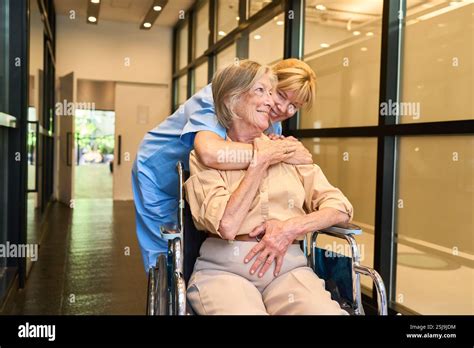 A Nurse Warmly Hugs A Senior Woman Seated In A Wheelchair Creating A Touching Moment Reflecting