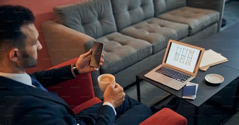 검은 양복을 입은 남자가 공항 라운지 구역에서 커피 한 잔을 즐기고 스마트폰 화면을 보고 있다 커피 테이블에 노트북 사진 Unsplash의 성공 이미지