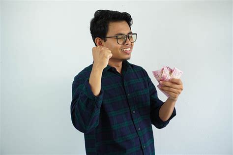 젊은 아시아 남자는 인도네시아 지폐를 들고 흰색 배경에 고립된 흥분된 표정을 보여주면서 주먹을 꽉 쥐었다 25 29세에 대한