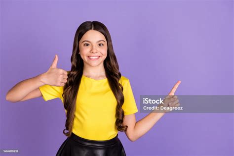 멋진 낙관적 인 갈색 머리 머리 소녀의 초상화는 생생한 라일락 색 배경에 고립 된 노란색 티셔츠를 입고 엄지 손가락 위로 포인트