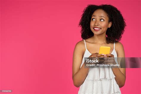 분홍색 배경에 고립 된 여유 공간을 보고있는 전화를 가진 행복한 사려 깊은 밀레니엄 아프리카 계 미국인 여성 여자에 대한 스톡