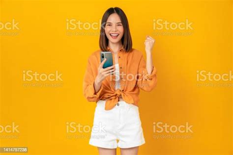 헤드폰을 들고 주먹으로 스마트폰을 들고 노란 배경에서 성공을 거둔 승리를 축하하는 쾌활한 젊은 아시아 여성 스튜디오 촬영에 대한