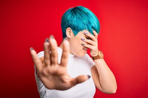 파란색 패션 머리를 가진 젊은 아름다운 여자는 손으로 눈을 덮고 슬프고 두려움 표정으로 중지 제스처를하고 빨간 배경 위에 캐주얼