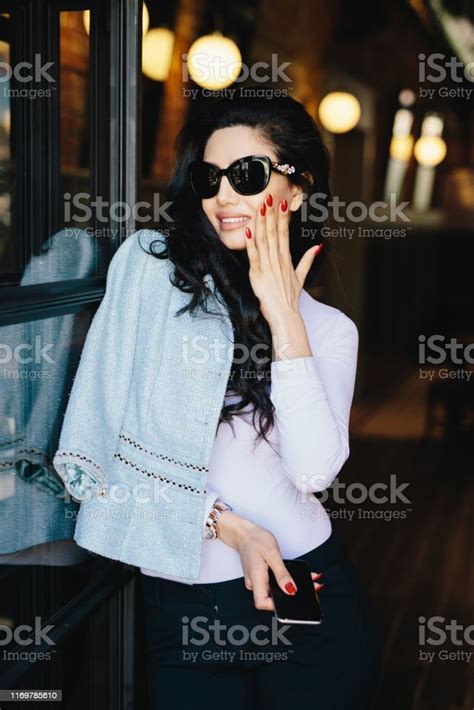 카페에 서있는 동안 그녀의 빨간 좋은 손톱을 보여주는 뺨에 손을 들고 선글라스와 정장 옷을 입고 어두운 고급스러운 머리를 가진 매력적인 사업가의 수직 초상화 갈색 머리에