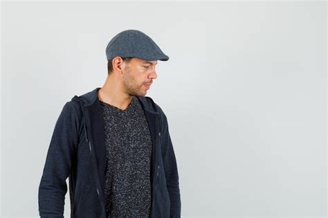 Молодой человек в футболке куртке кепке смотрит вниз и задумчиво вид спереди Бесплатно Фото