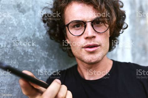 근접 촬영 야외 서 하 고 휴대 전화에 젊은 남자의 초상화 젊은 남자 곱슬 머리와 구체적인 배경에 그의 휴대 전화에 전화를 만드는 밖으로 휴식 하는 둥근 안경 착용 거리에