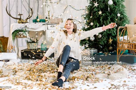 청바지와 니트 흰색 스웨터에 예쁜 젊은 여자는 크리스마스 트리 근처에 앉아 황금 색종이를 던져 메리 크리스마스 새해 가족 휴일의 개념입니다 12월에 대한 스톡 사진 및 기타