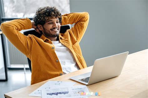 세련된 캐주얼 옷을 입은 긍정적 인 인도 남자 회사 직원은 업무용 책상에 앉아 노트북으로 일하면서 휴식을 취하고 머리 뒤로 손을