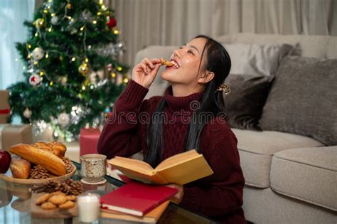 A Cheerful Asian Woman Enjoys Christmas Cookies And A Warm Drink While Reading In Her Living