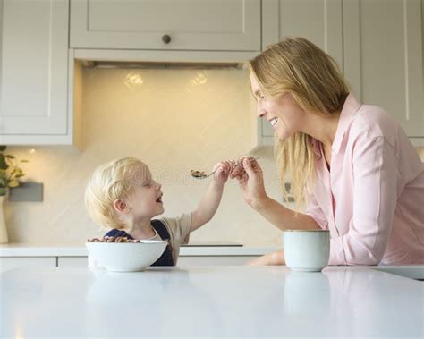 Mother At Home Encouraging Young Son To Eat Breakfast In Kitchen Stock