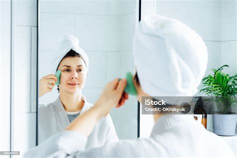 목욕 가운을 입은 여성은 욕실에서 구아 샤 롤러로 얼굴 마사지를하는 그녀의 머리에 수건을 들고 있습니다 스킨 케어에 대한 천연 절차의 개념 홈 뷰티 셀프 케어 트리트먼트