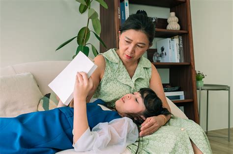 교복을 입은 어린 동양인 딸이 엄마 무릎에 누워 오후에 학교에서 돌아온 후 함께 책을 읽고 있습니다 함께 시간을 보내는 행복한