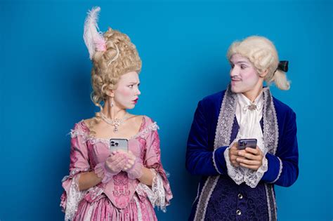 바로크 양식의 의상을 입은 기발한 커플이 파란색 배경에서 스마트폰으로 문자를 보내며 과거의 우아함과 현대 기술이 조화를 이루고 있습니다 16 17 살에 대한 스톡 사진 및
