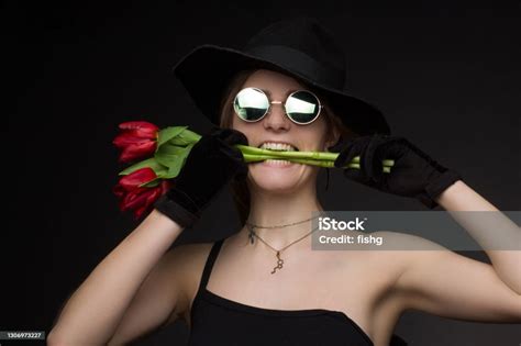 모자와 선글라스를 입은 젊은 여성의 스튜디오 초상화가 그녀의 입에 빨간 튤립 꽃다발을 들고 검은 색 배경에 검은색에 대한 스톡 사진 및 기타 이미지 Istock