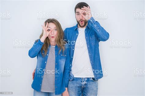 흰색 고립 된 배경 위에 함께 서있는 젊은 아름다운 부부는 손가락을 통해 보고 놀란 얼굴 눈으로 충격을 받은 확인 제스처를하고