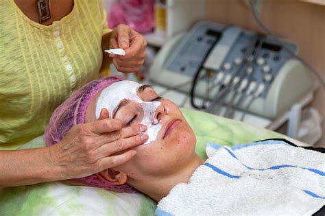 숙련 된 미용 학자는 얼굴 클렌징 및 미용 치료 중에 소파에 놓여 있는 어린 소녀의 얼굴에 흰 크림 마스크를 적용 합니다 내각